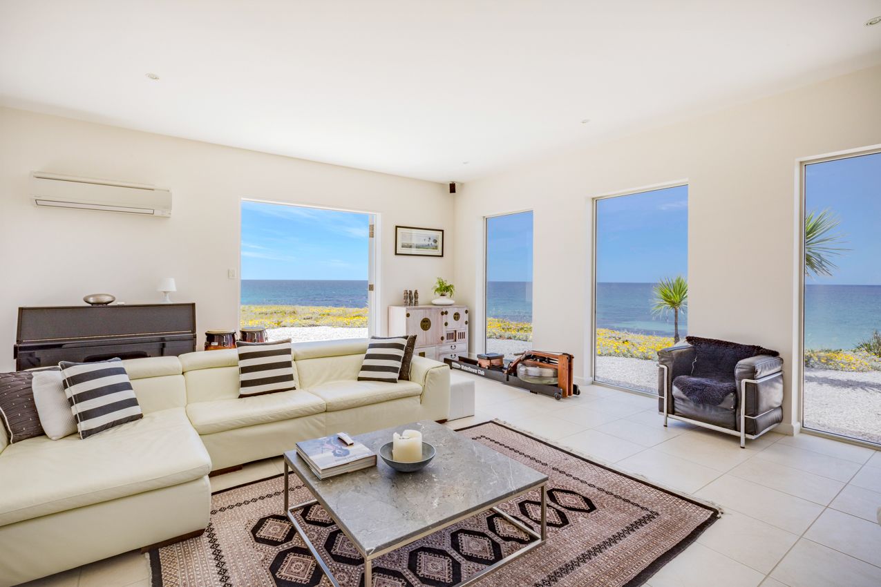 Uma sala de estar ampla, com um sofá grande, uma mesa de centro e uma televisão. Janelas grandes oferecem vista para a praia.