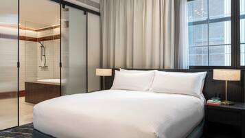 Quarto de hotel com uma cama grande, mesa de cabeceira com abajur, janela com cortinas e vista para a cidade.