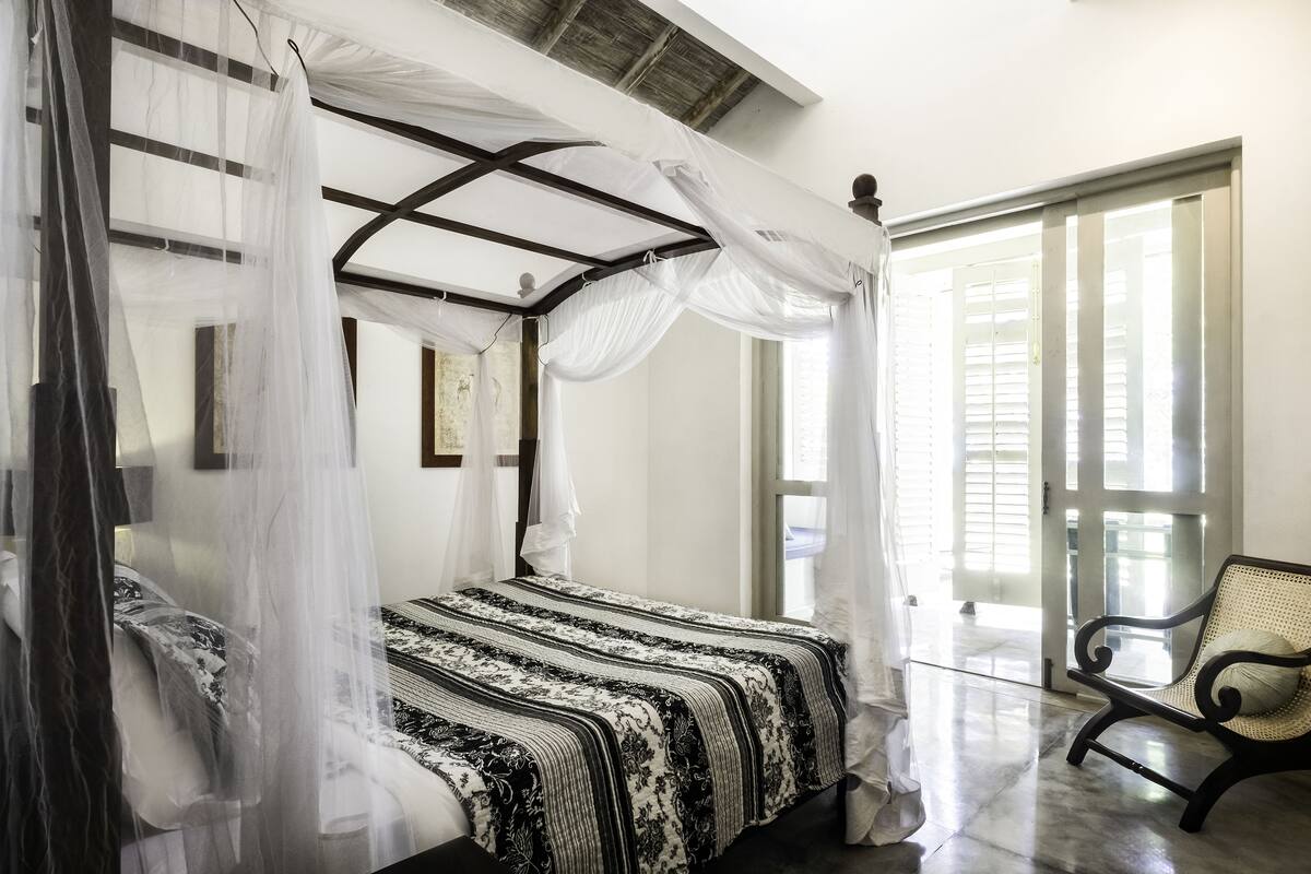 Uma cama de dossel com colcha estampada, uma cadeira e vista exterior por uma porta de vidro.