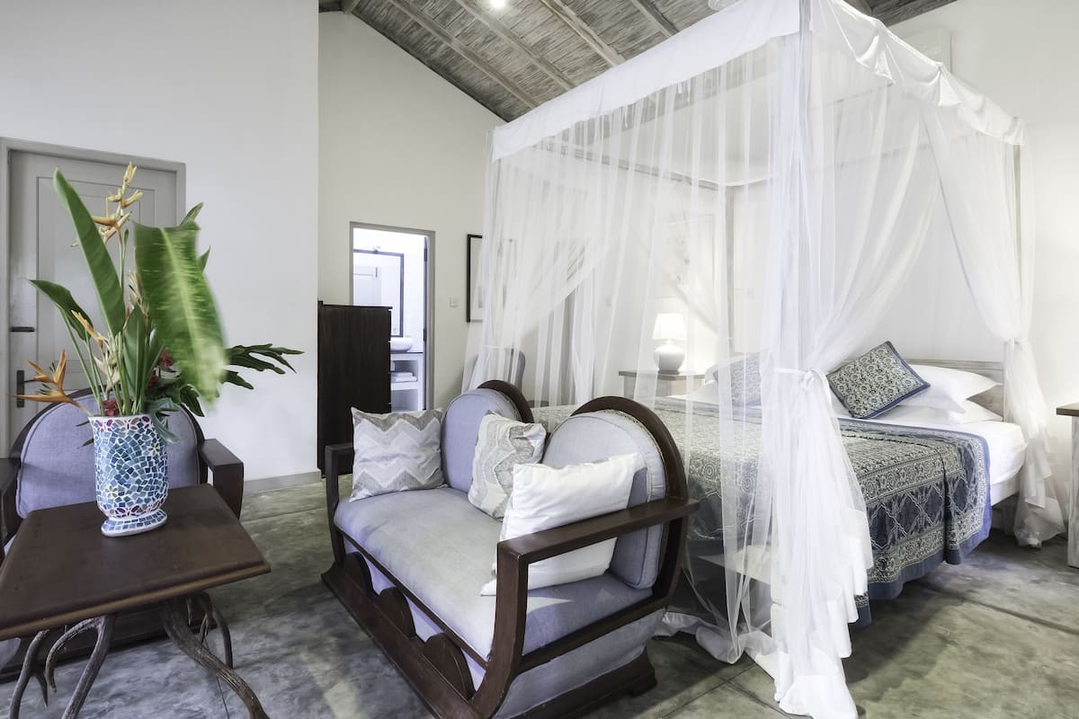 Quarto com cama de dossel, sofá, mesinha com planta e vista para o exterior.