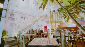 Um restaurante com mesas e cadeiras de madeira, uma escadaria e obras de arte nas paredes.