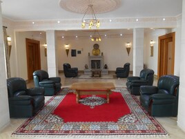 Uma sala de estar ampla com uma mesa de centro, rodeada por poltronas de couro, um tapete estampado em vermelho e uma lareira com objetos decorativos.