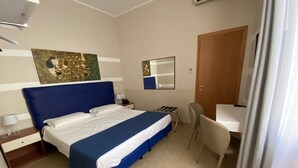 Quarto de hotel com uma cama grande, uma escrivaninha, uma cadeira, uma mesinha de cabeceira, um espelho e obras de arte na parede.