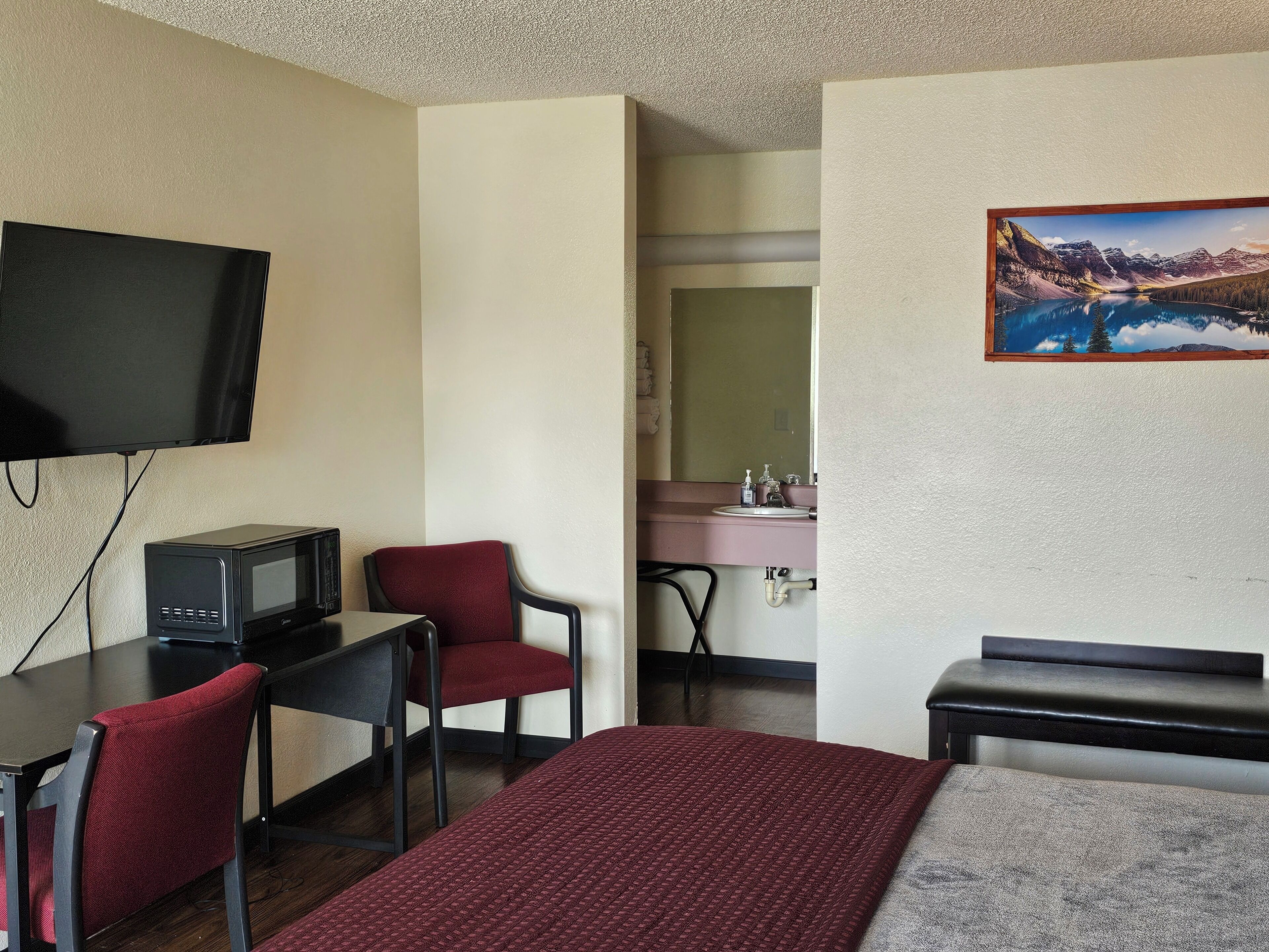 Quarto de hotel com uma cama, uma escrivaninha com televisão, uma cadeira, uma pia e um quadro de uma paisagem montanhosa.