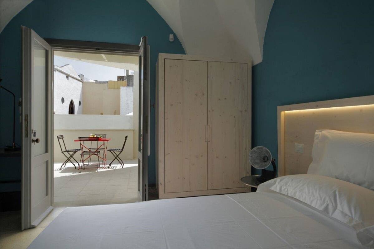 Quarto com cama, guarda-roupa, mesa e cadeiras, e vista para área externa por uma porta aberta.