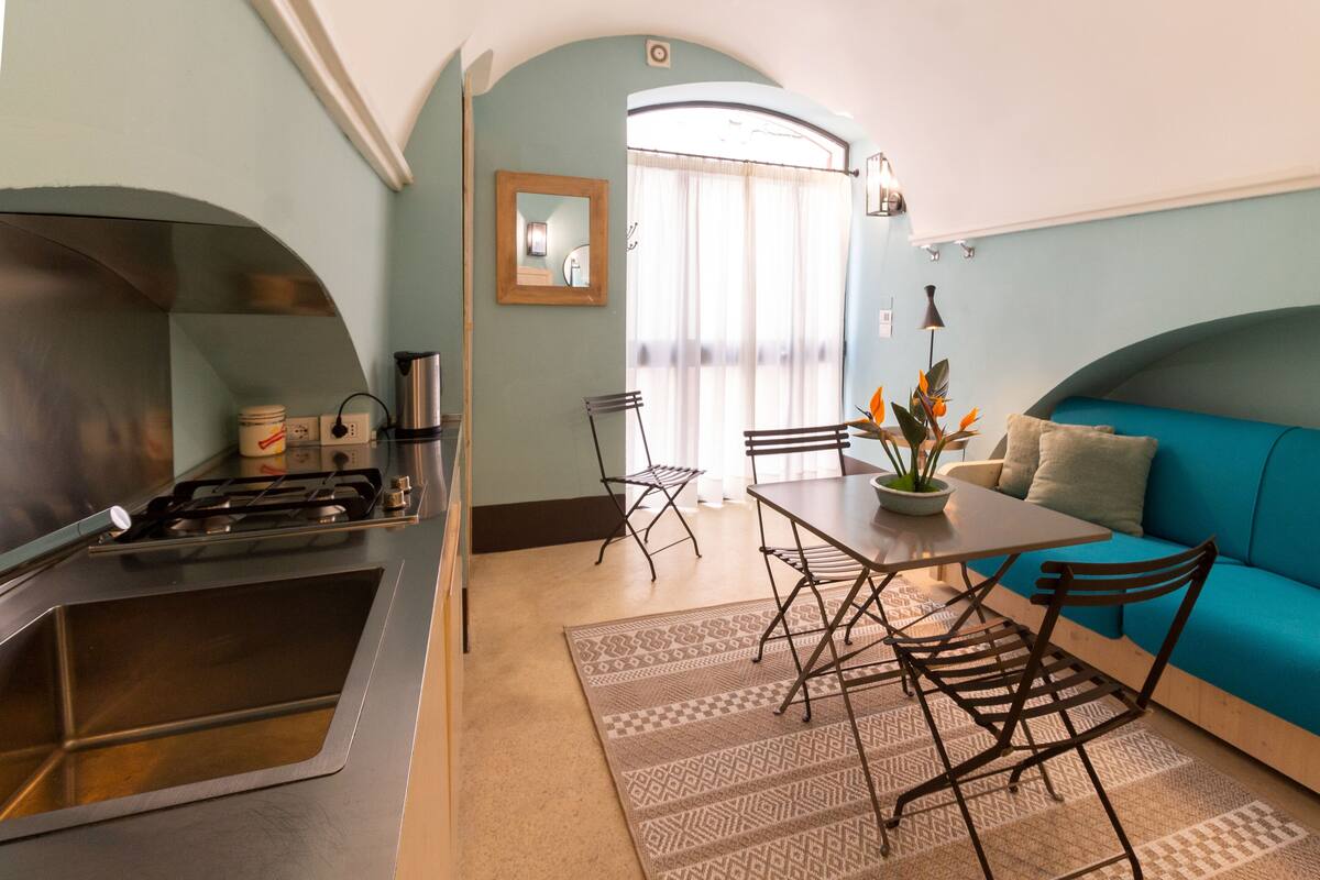 Cozinha compacta com fogão a gás, pia e área de refeições com mesa e cadeiras. Há um sofá azul e uma janela com cortinas na sala.