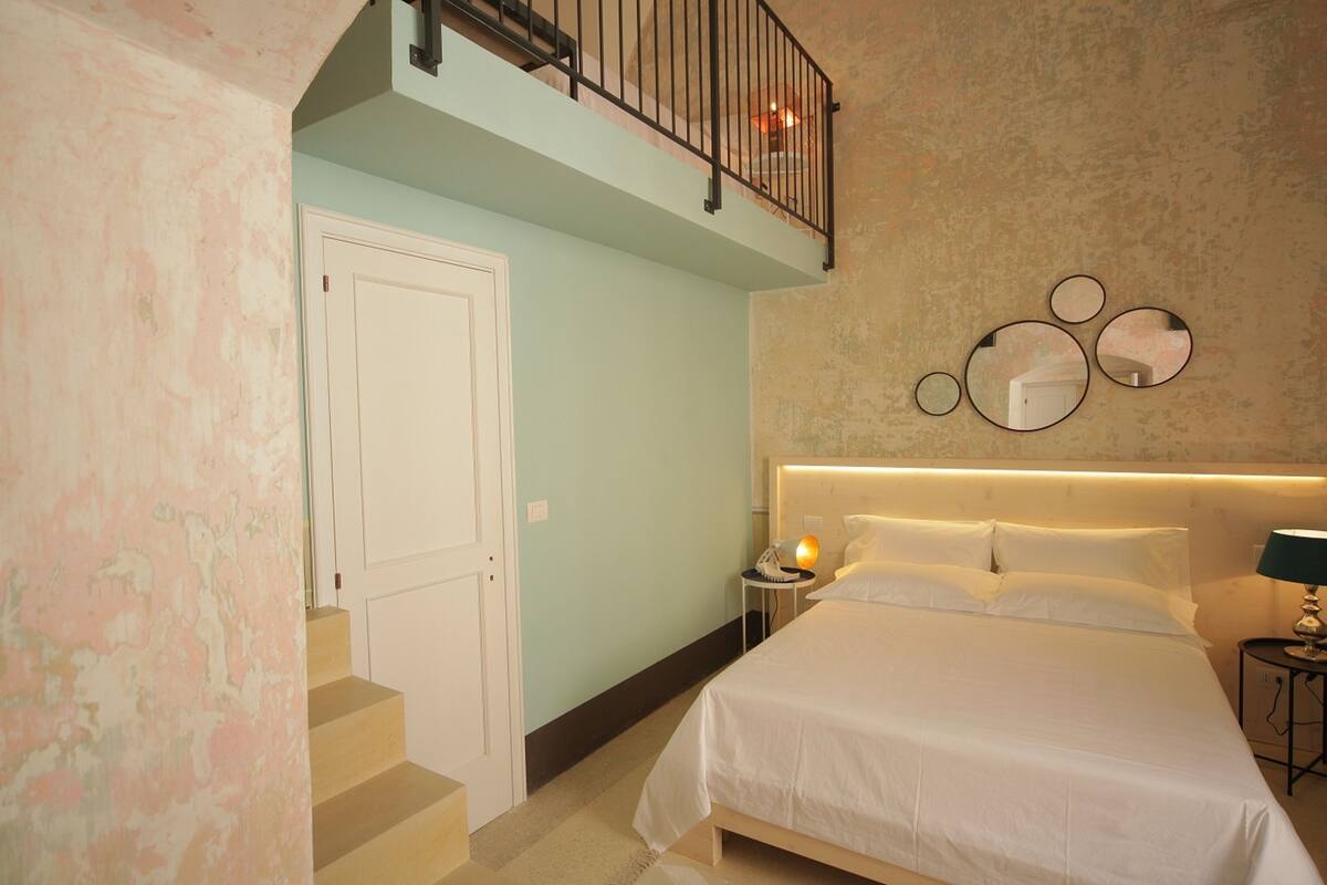 Quarto com cama, mesa de cabeceira, espelho fixado na parede e escada que leva a um andar superior.