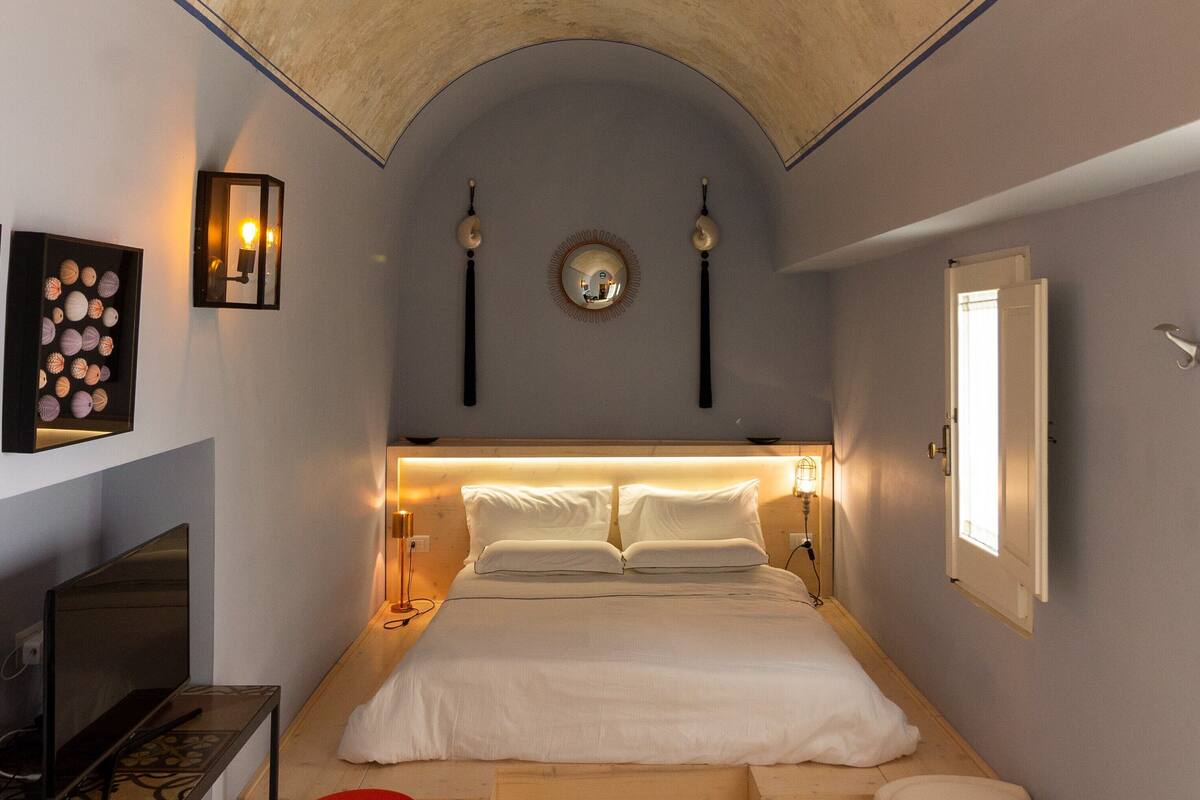 Quarto com cama, abajures, espelho, TV na parede e um banquinho vermelho pequeno.