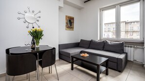 Uma sala de estar moderna com um sofá cinzento, uma mesa de centro preta e uma mesa de jantar com cadeiras. Há uma janela com vista para edifícios.