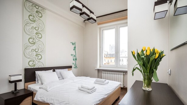 Quarto moderno com cama de madeira, roupa de cama branca, mesa de cabeceira com abajur, janela com vista para edifícios e um vaso com flores amarelas em uma cômoda.