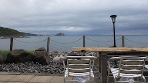 Vista para o mar com uma mesa de madeira e duas cadeiras de metal, um poste de luz e uma barreira de corda.