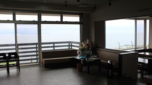 Quarto com janelas amplas com vista para a água, área de estar com um banco e uma mesa pequena com um vaso de flores.