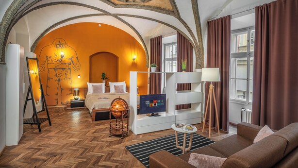 Quarto de hotel com soalho de madeira em padrão herringbone, cama com cabeceira, televisão, secretária, candeeiro, sofá e uma grande janela com cortinas.
