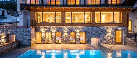 Hotel de dois andares com piscina, fachada de pedra e varandas iluminadas.