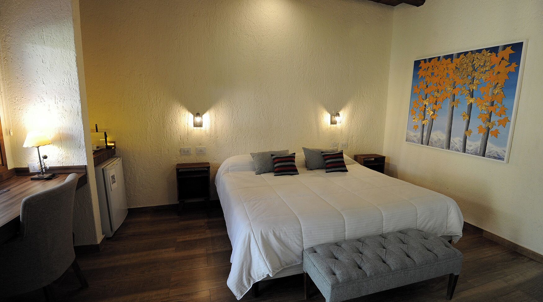 Quarto com cama, mesas de cabeceira, escrivaninha, luminária e um quadro na parede.