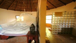 Quarto com telhado de palha, cama com mosquiteiro, armário de madeira e janela com vista para o exterior.