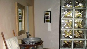 Um quarto com piso de azulejos, uma cadeira de madeira com tecido branco, uma bacia de metal, um espelho e uma vitrine de vidro com objetos.