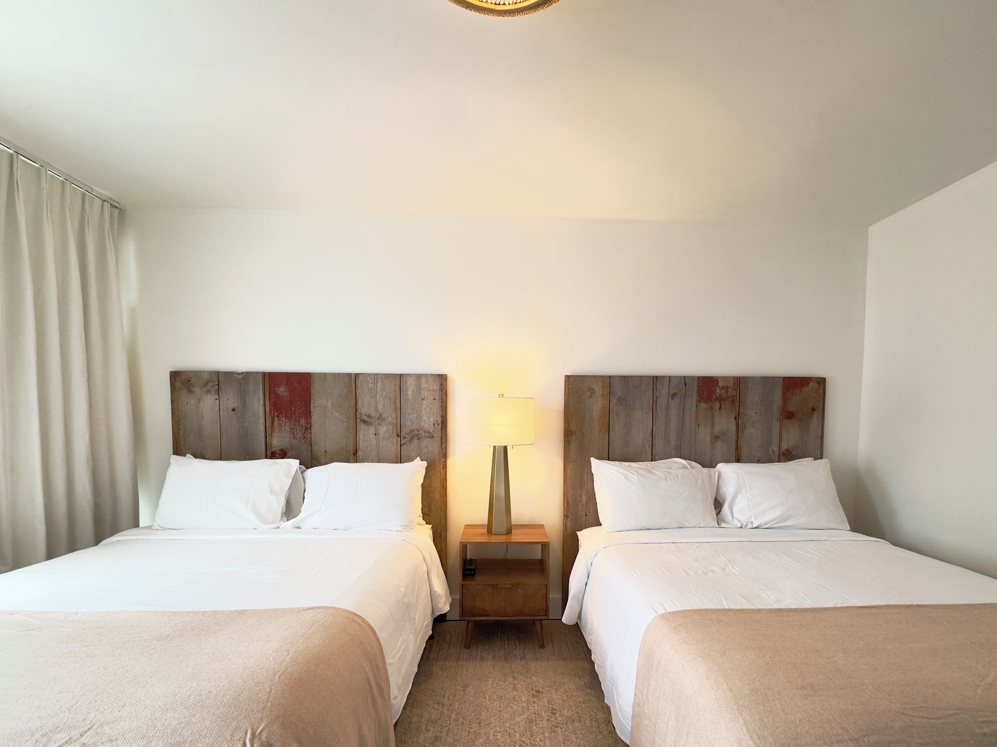Quarto de hotel com duas camas, uma mesa de cabeceira com abajur e uma janela com cortinas.