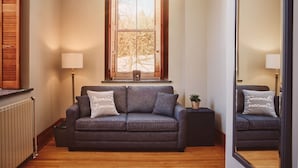 Uma sala de estar acolhedora com um sofá cinzento, duas almofadas iguais, uma mesinha lateral, um candeeiro de pé e uma janela com vista para árvores.