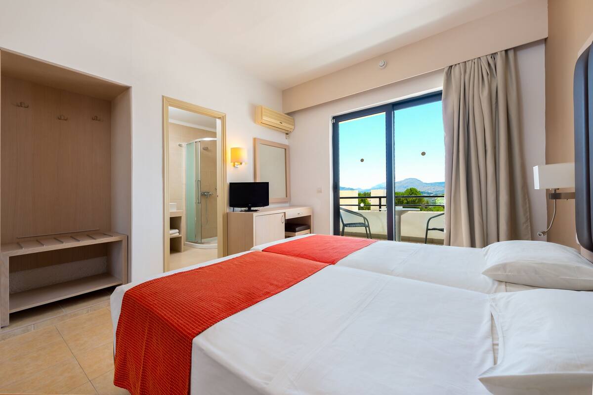 Quarto de hotel com cama, escrivaninha, televisão e varanda com vista para as montanhas.