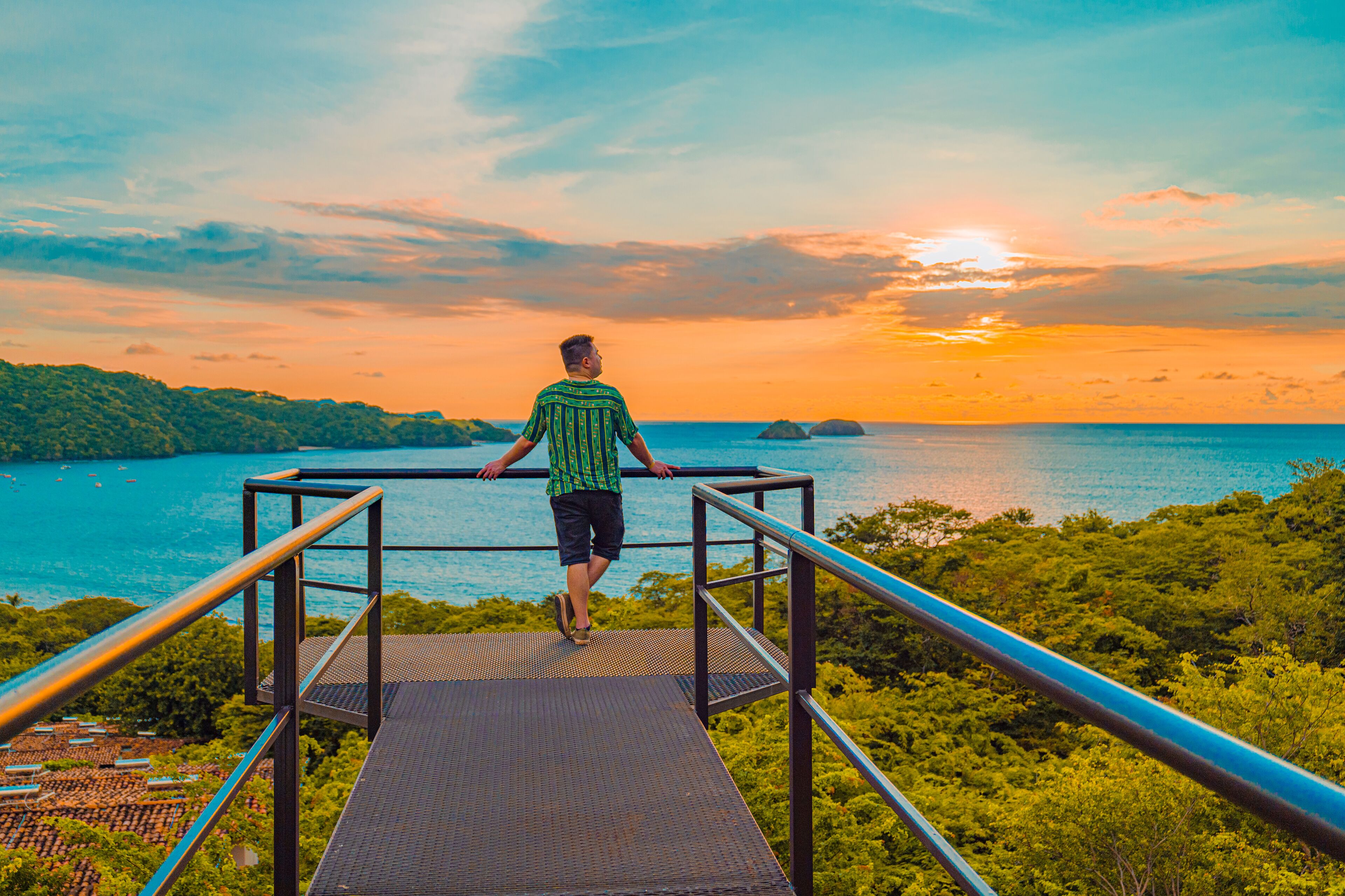 Um homem está em uma plataforma de metal com grades, apreciando a paisagem costeira com árvores e o mar.