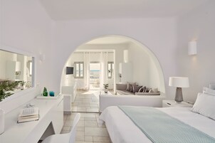 Quarto de hotel moderno com uma cama grande, uma escrivaninha, uma cadeira e vista para o mar através de uma porta em arco.