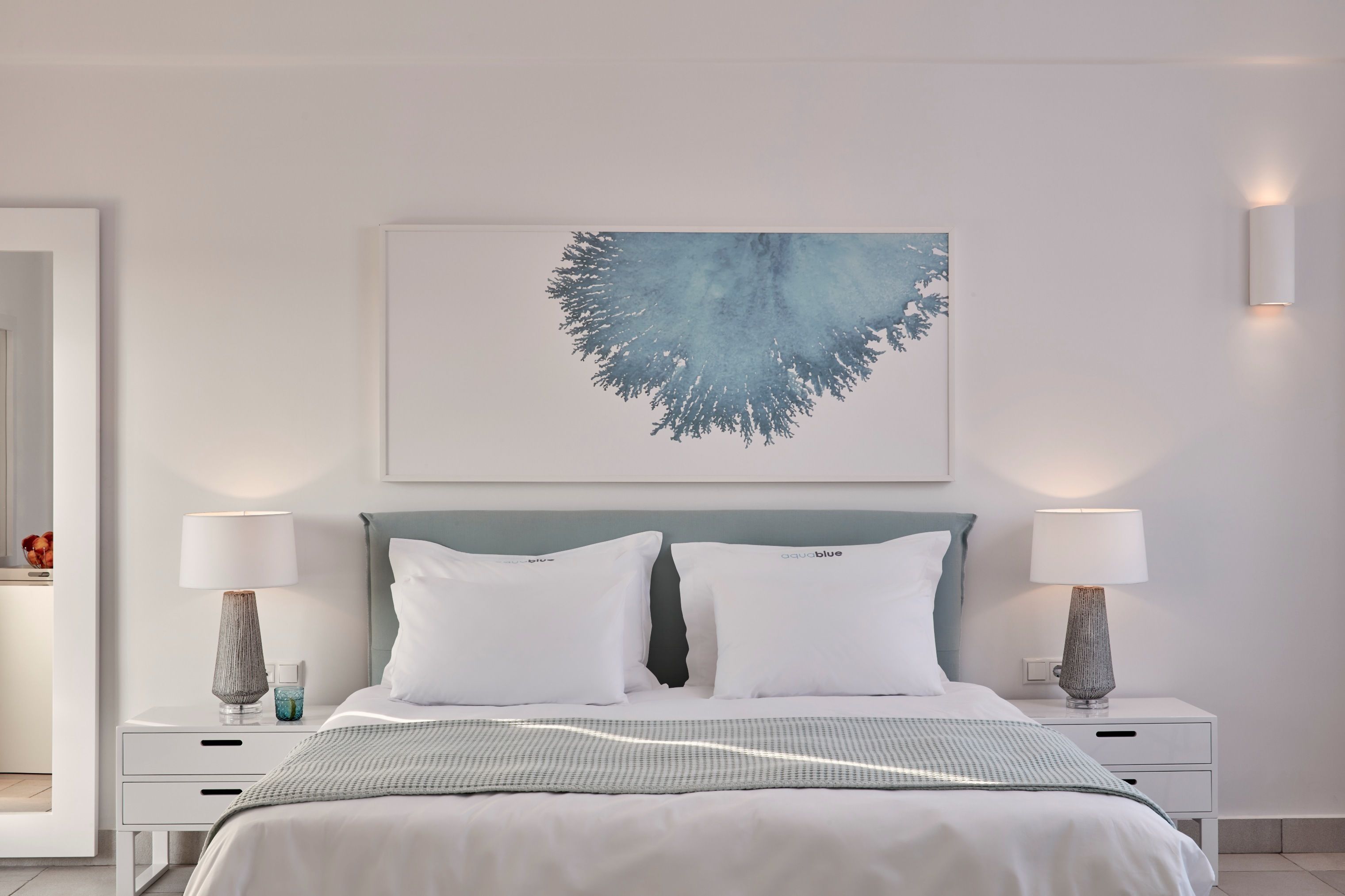 Quarto moderno com cama, duas mesas de cabeceira, um criado-mudo, uma luminária na parede e um quadro abstrato grande.