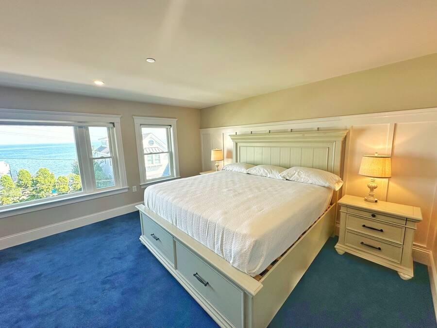 Quarto com cama grande, duas mesas de cabeceira e vista para o mar através de duas janelas.