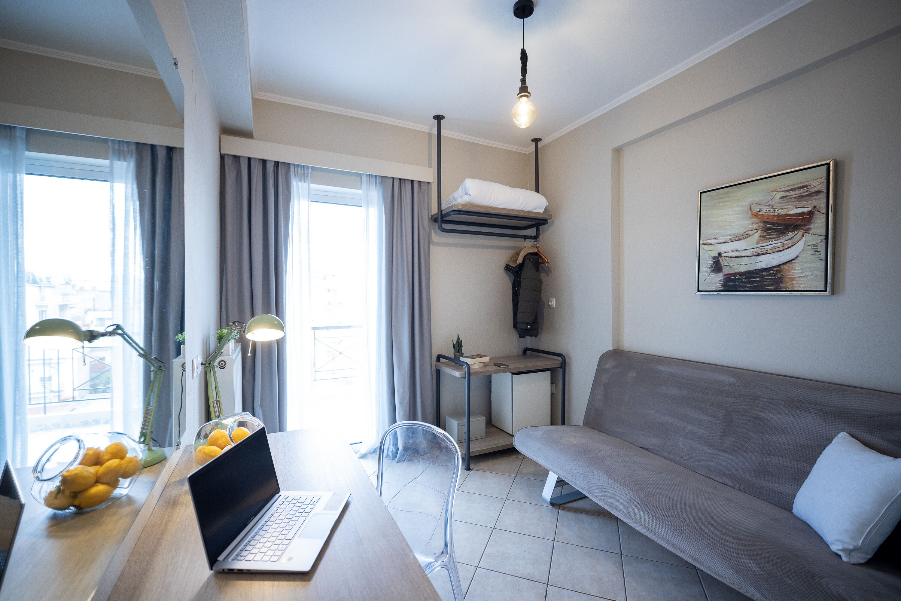 Uma sala de estar moderna com um sofá, uma escrivaninha com um laptop, uma cadeira transparente, um abajur e um quadro na parede.