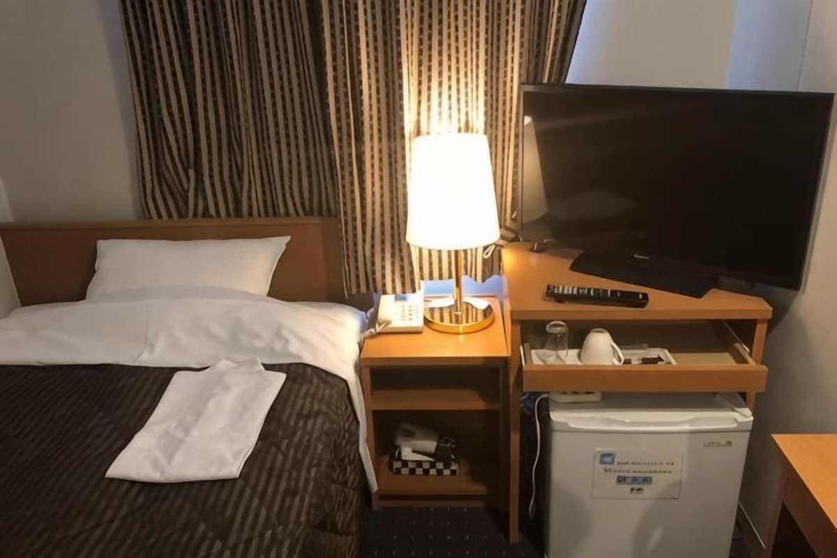 Quarto de hotel com cama, televisão, escrivaninha com luminária, minibar e cortina.