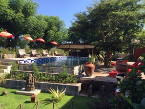 Uma área de piscina com espreguiçadeiras, guarda-sóis vermelhos e uma piscina revestida em azulejos.