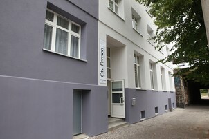 Um edifício com fachada cinza e branca, uma porta com uma placa e uma árvore ao primeiro plano.