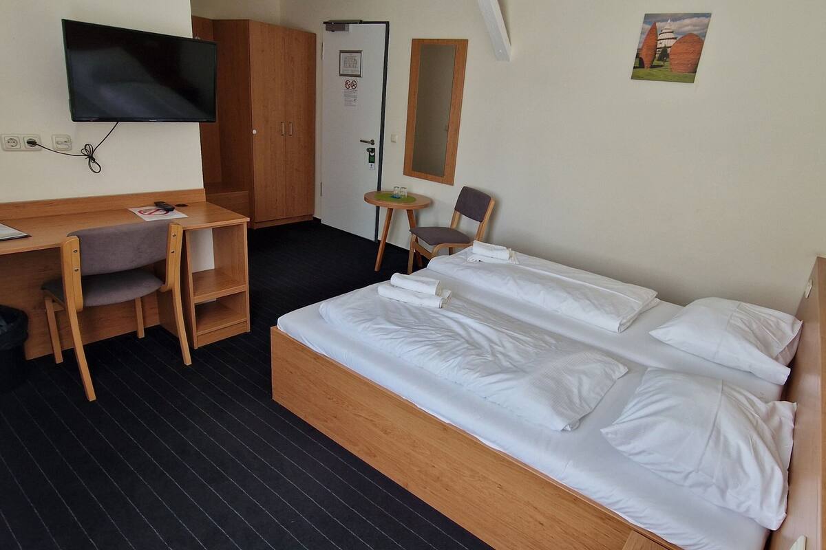 Quarto de hotel com cama, escrivaninha, cadeira, televisão e uma pequena mesa com uma cadeira.