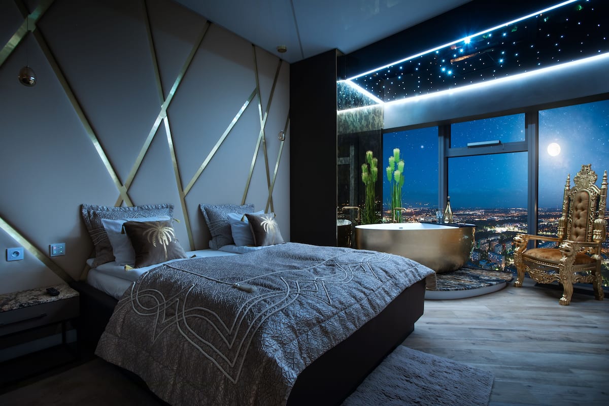 Quarto com uma cama grande, banheira, poltrona dourada e vista para a cidade à noite.