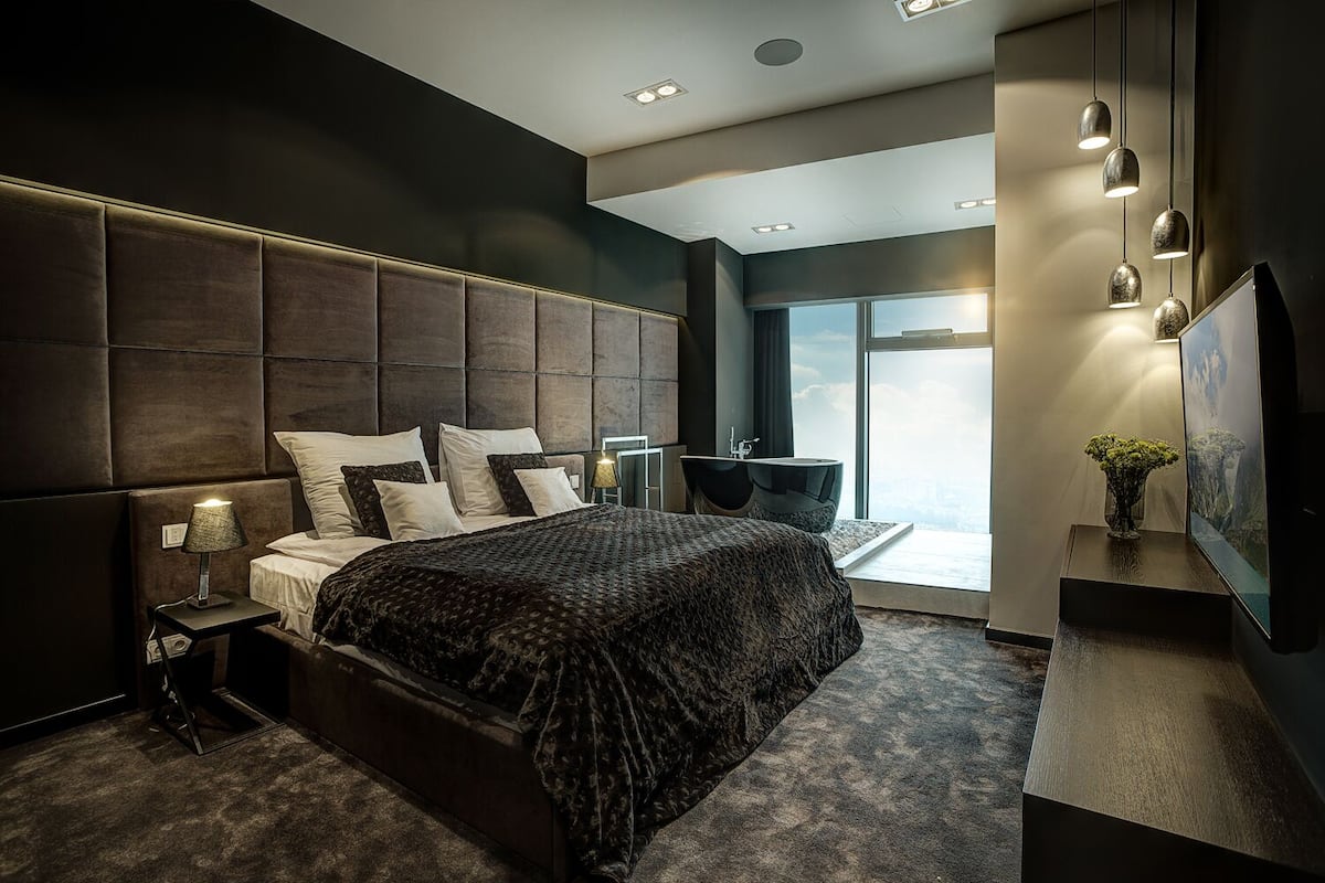 Quarto moderno com cama grande, mesa de cabeceira, penteadeira e uma janela ampla com vista para a cidade.