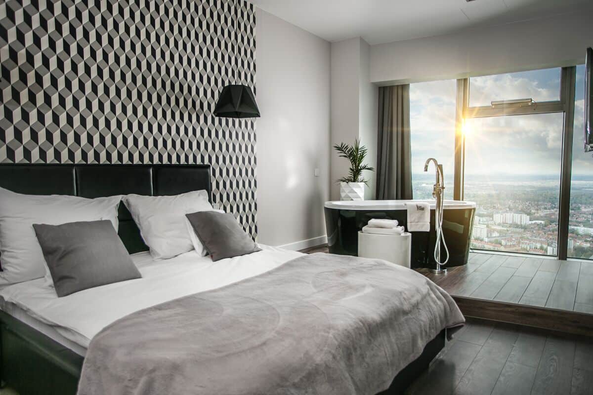 Quarto de hotel moderno com uma cama grande, parede com padrão geométrico, vista da cidade por uma janela ampla e banheiro pequeno com pia e suporte para toalhas.
