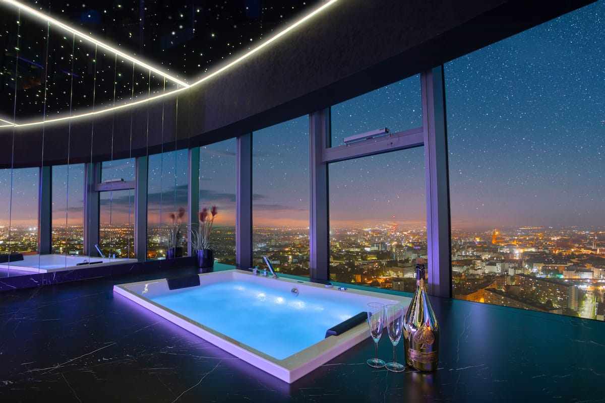 Um spa moderno com uma banheira grande, vista da cidade através de amplas janelas, e céu estrelado visível pela janela.