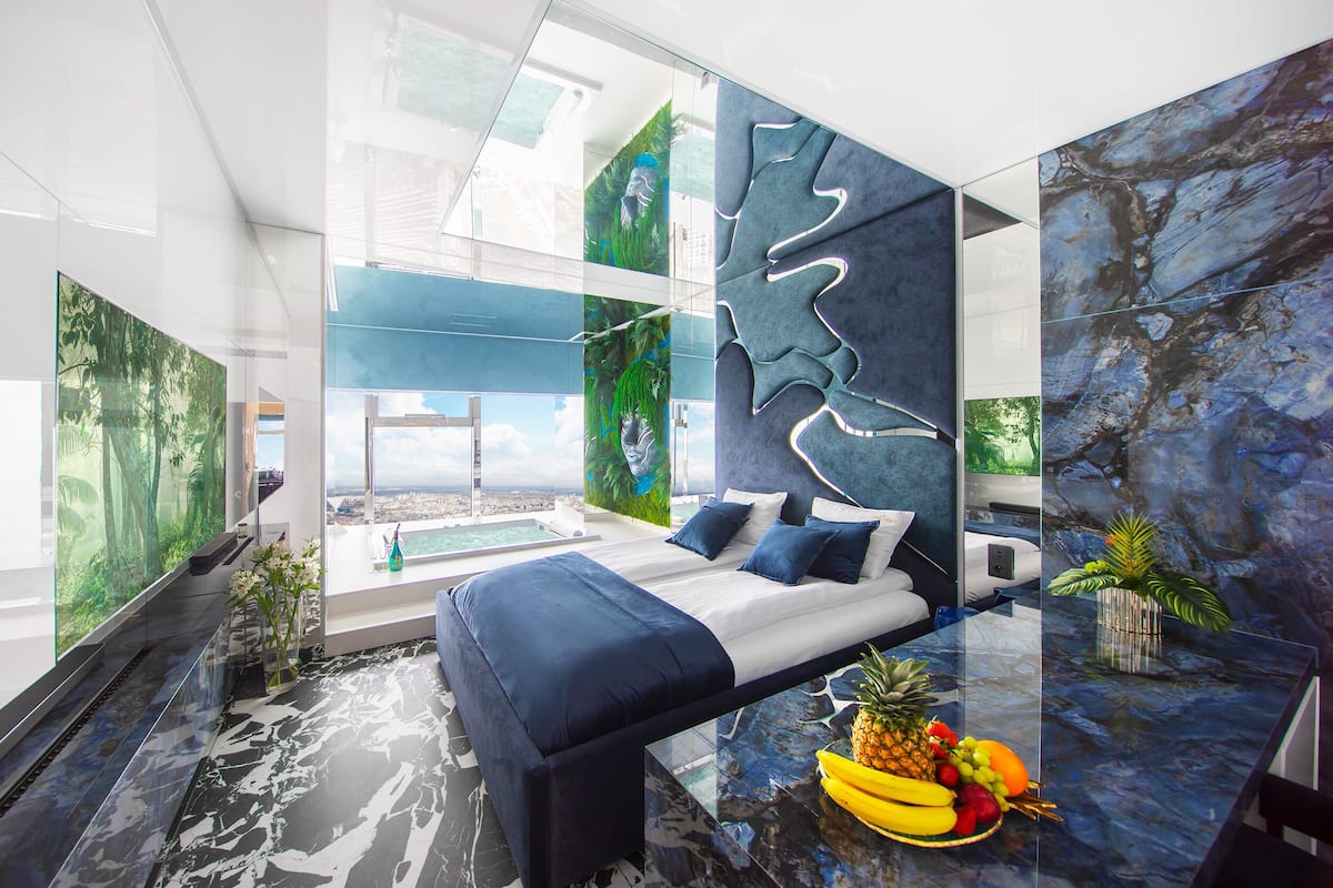 Quarto moderno com cama de vidro, vista para a cidade e um mural tropical.