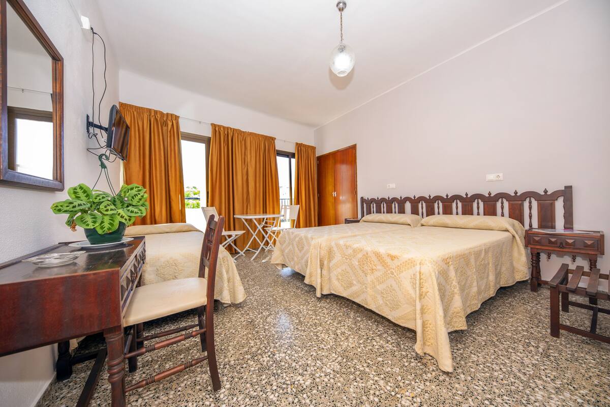Quarto de hotel com cama, escrivaninha, cadeira, mesa, planta em vaso e janela com cortinas.