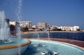 Uma fonte à beira-mar com vários jatos de água, rodeada por uma piscina. Ao fundo, vários edifícios altos com varandas, provavelmente hotéis ou apartamentos.