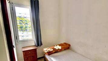 Quarto pequeno com cama de solteiro, piso de azulejos, mesa de cabeceira de madeira, janela com cortinas e vista para o verde lá fora.