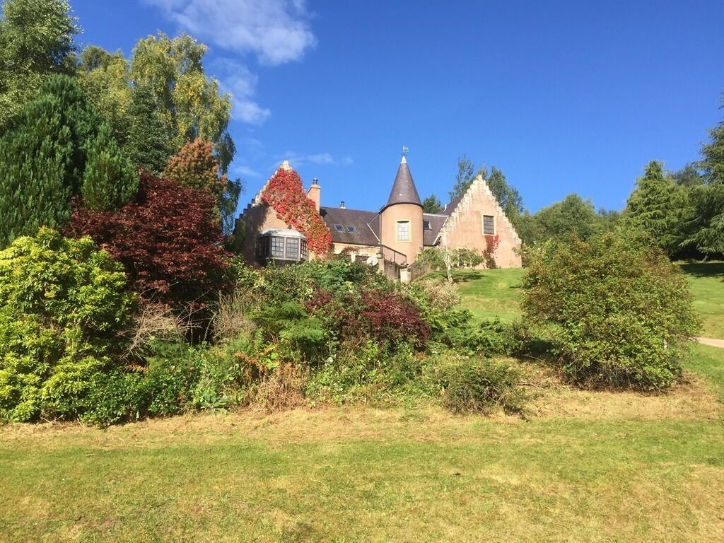 Uma casa grande e histórica, com uma torre imponente e várias chaminés, rodeada por vegetação exuberante e um gramado bem cuidado.