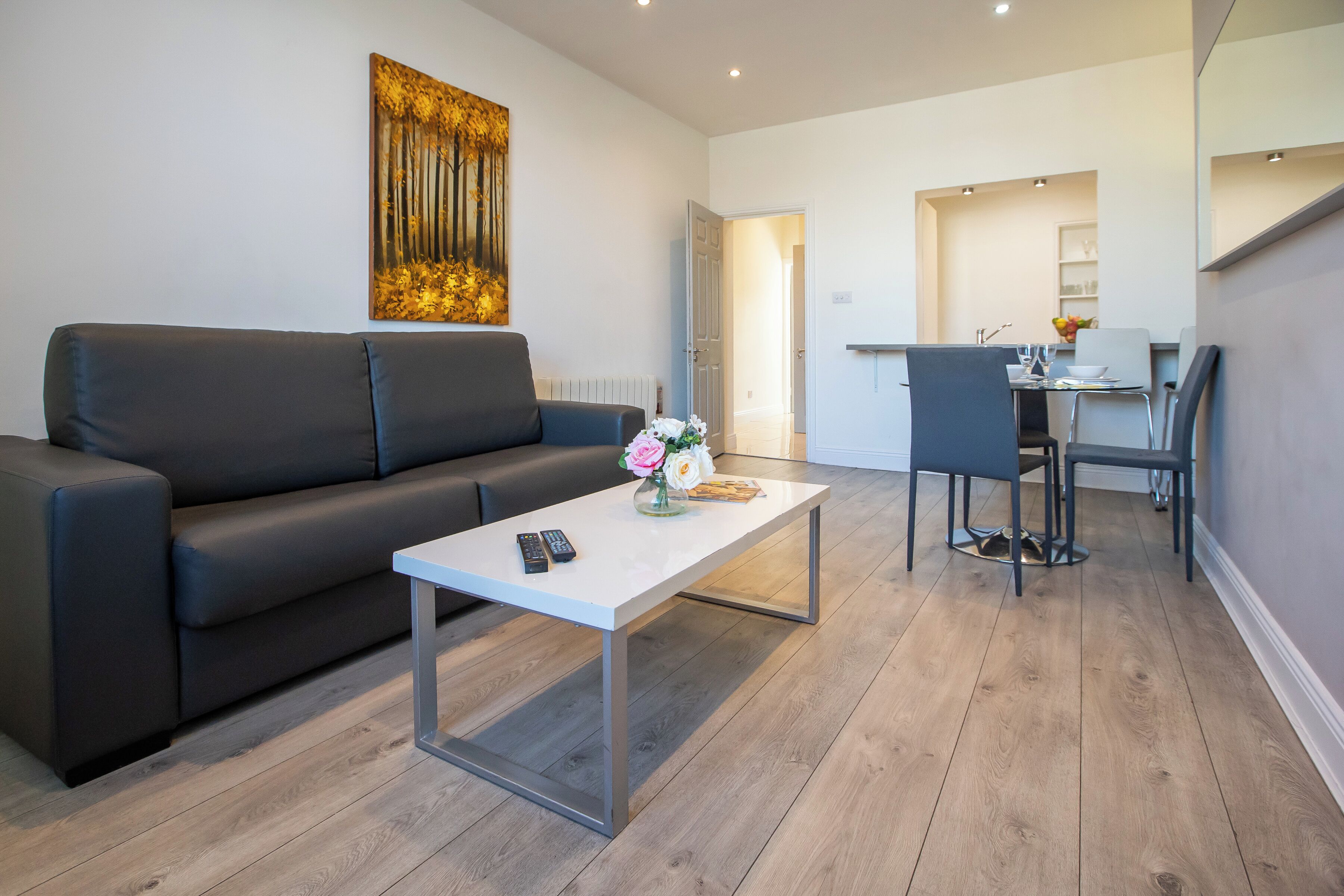 Uma sala de estar moderna com um sofá de couro preto, uma mesa de centro branca com um vaso de flores e um espaço de jantar com mesa e cadeiras.
