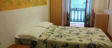 Quarto com cama, mesa de cabeceira de madeira e porta que dá para uma varanda.
