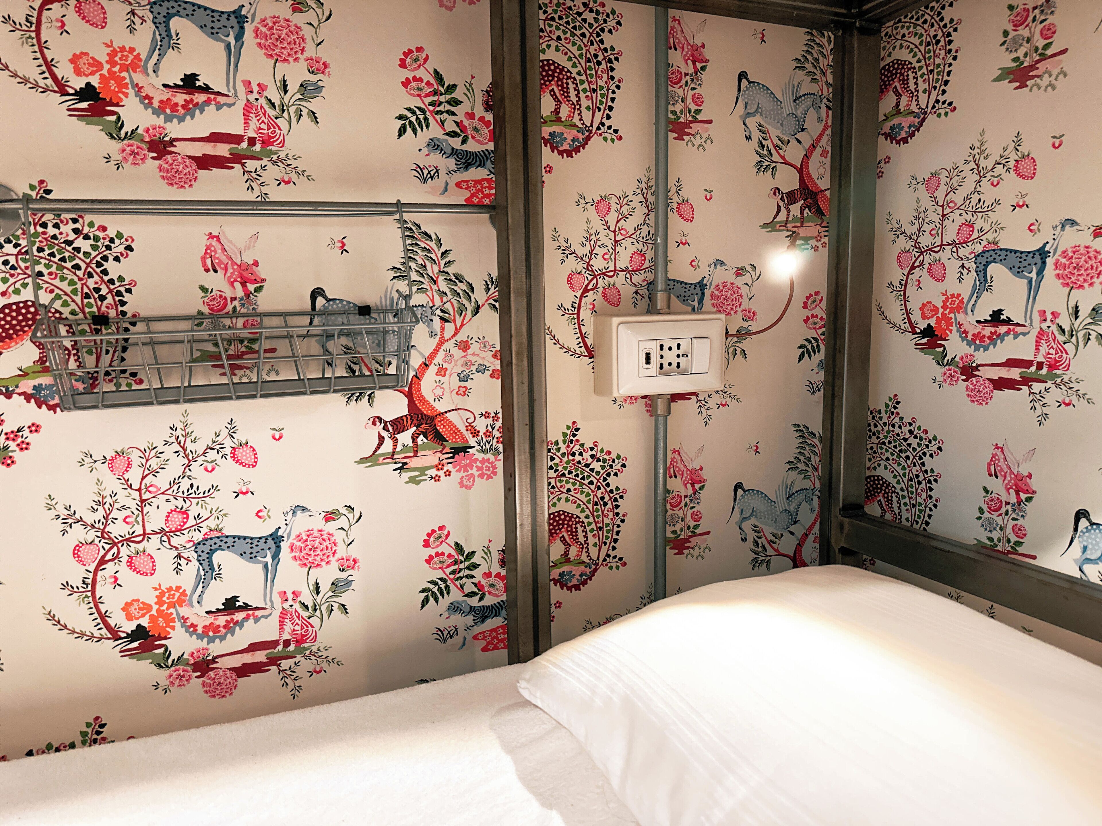 Quarto com papel de parede floral, uma cama, uma estante com objetos decorativos e uma tomada.