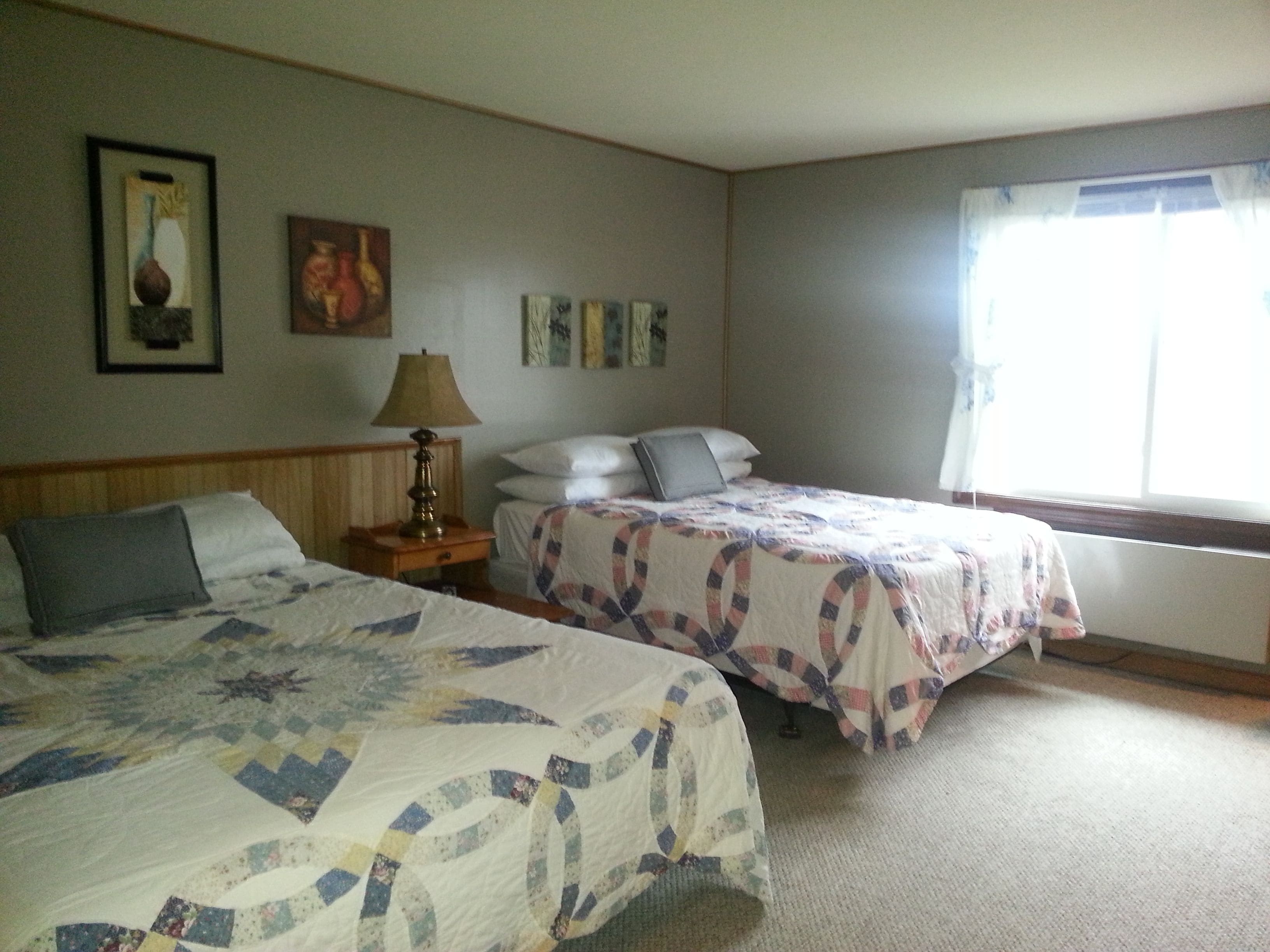 Quarto com duas camas, uma mesa de cabeceira com abajur e janela com cortinas.