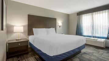 Quarto de hotel com uma cama grande, mesa de cabeceira com abajur, luz na parede e janela com cortinas.