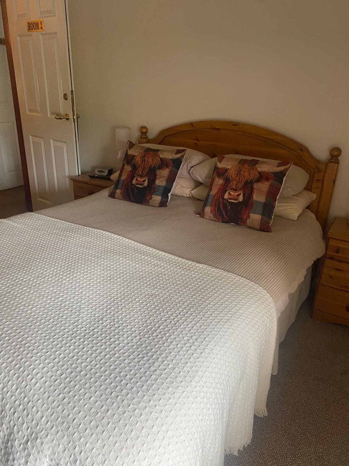 Cama com manta branca texturizada, duas almofadas com padrão xadrez, cabeceira de madeira e mesa de cabeceira com abajur.