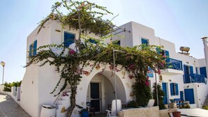Um edifício branco com janelas azuis e varanda, rodeado por vegetação e flores.