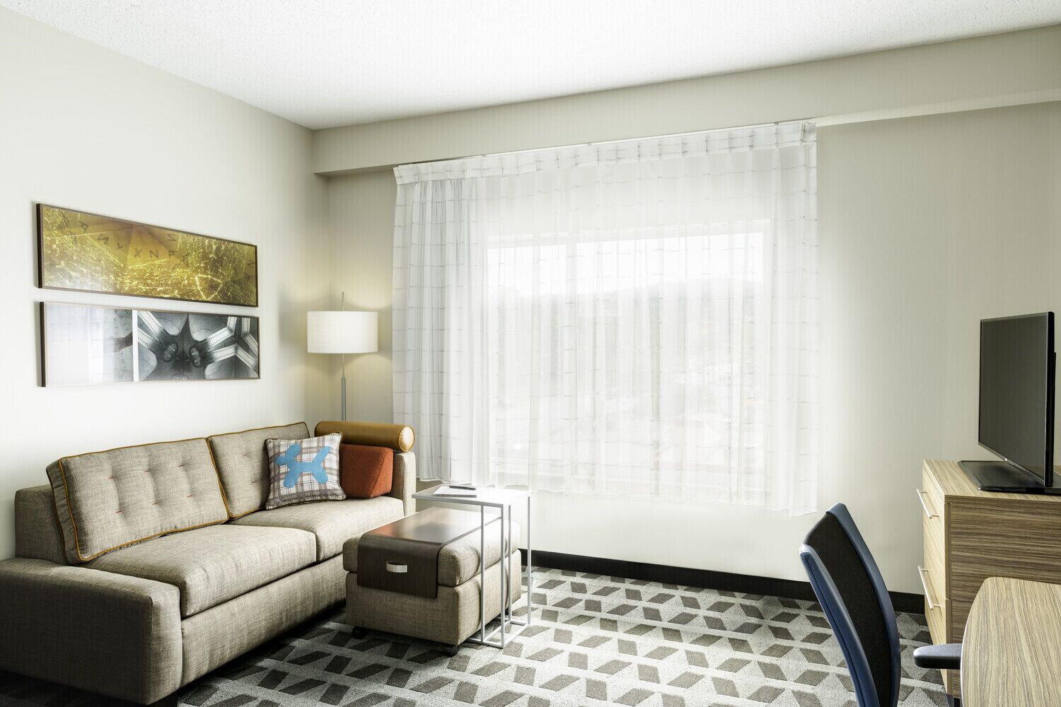 Quarto de hotel com sofá, mesinha, televisão e janela com cortinas leves.